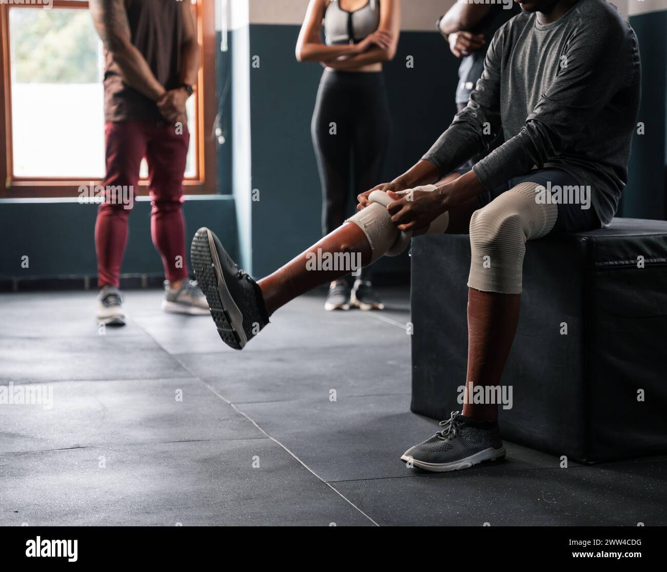 A man with a bandaged leg is seated on a gym bench surrounded by sports ...