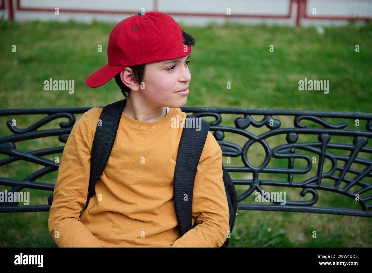 Portrait of a happy teenage schoolboy in a red cap, sitting on a park ...