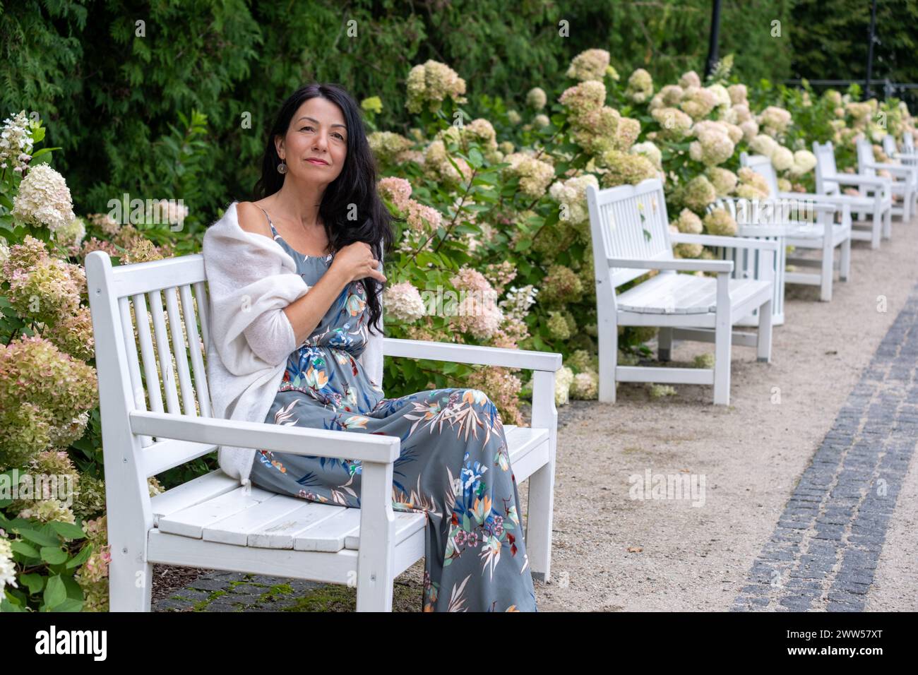 A pensive woman in a floral dress lounges on a white garden bench, her ...