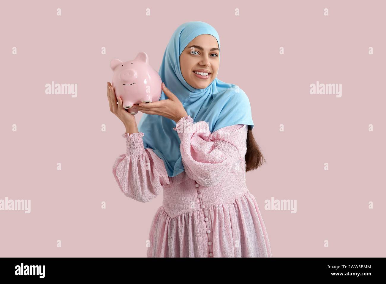 Beautiful young happy Muslim woman with piggy bank on pink background ...