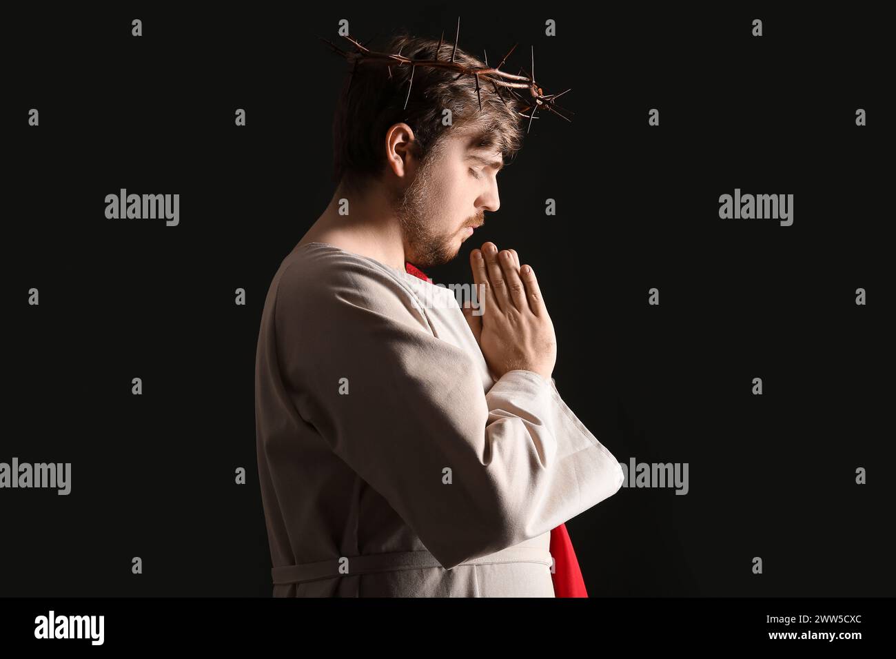 Man in Jesus robe and crown of thorns praying with light on black ...