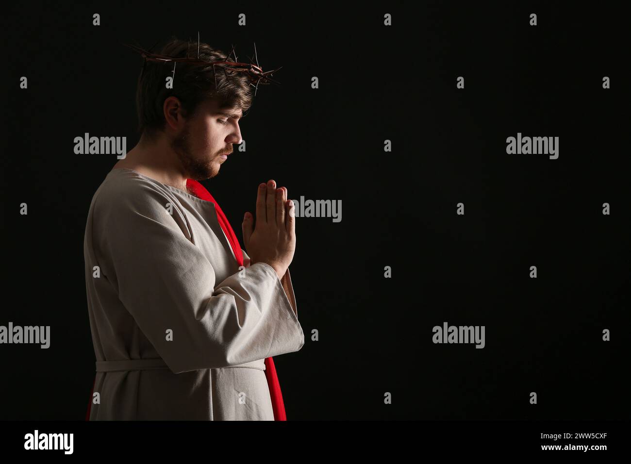 Man in Jesus robe and crown of thorns praying with light on black ...