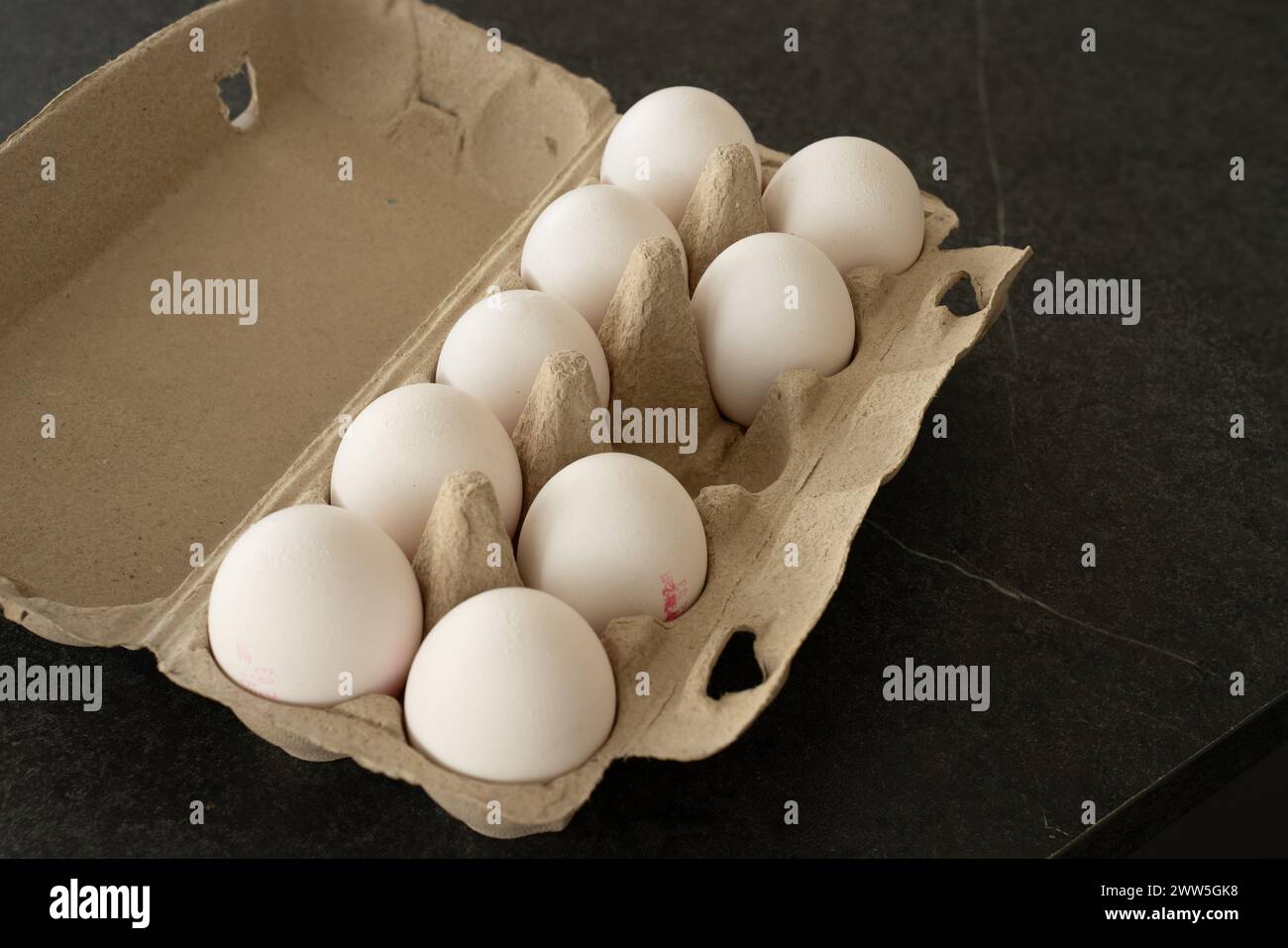 Brown open egg box on a dark table with nine white eggs in it. One egg ...