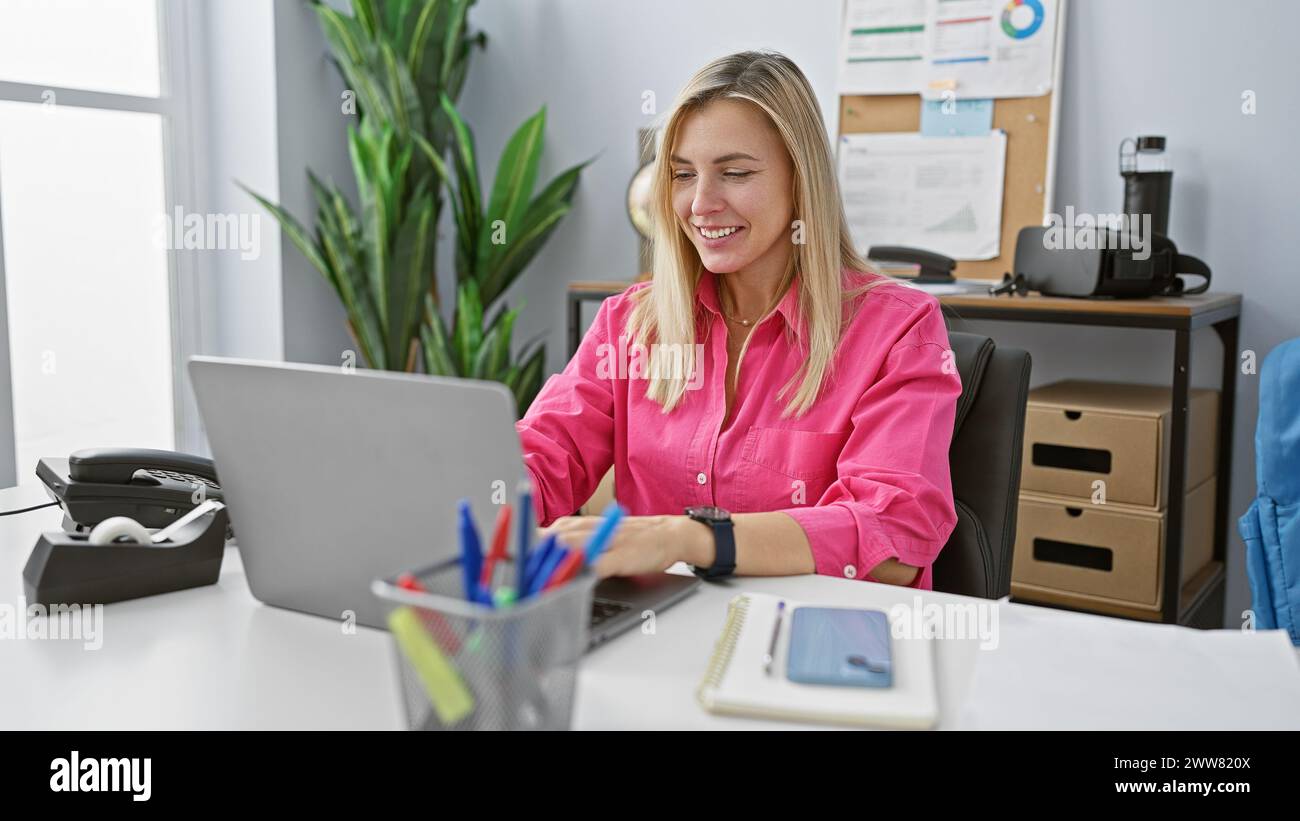 A smiling blonde woman working on a laptop in a modern office setting ...