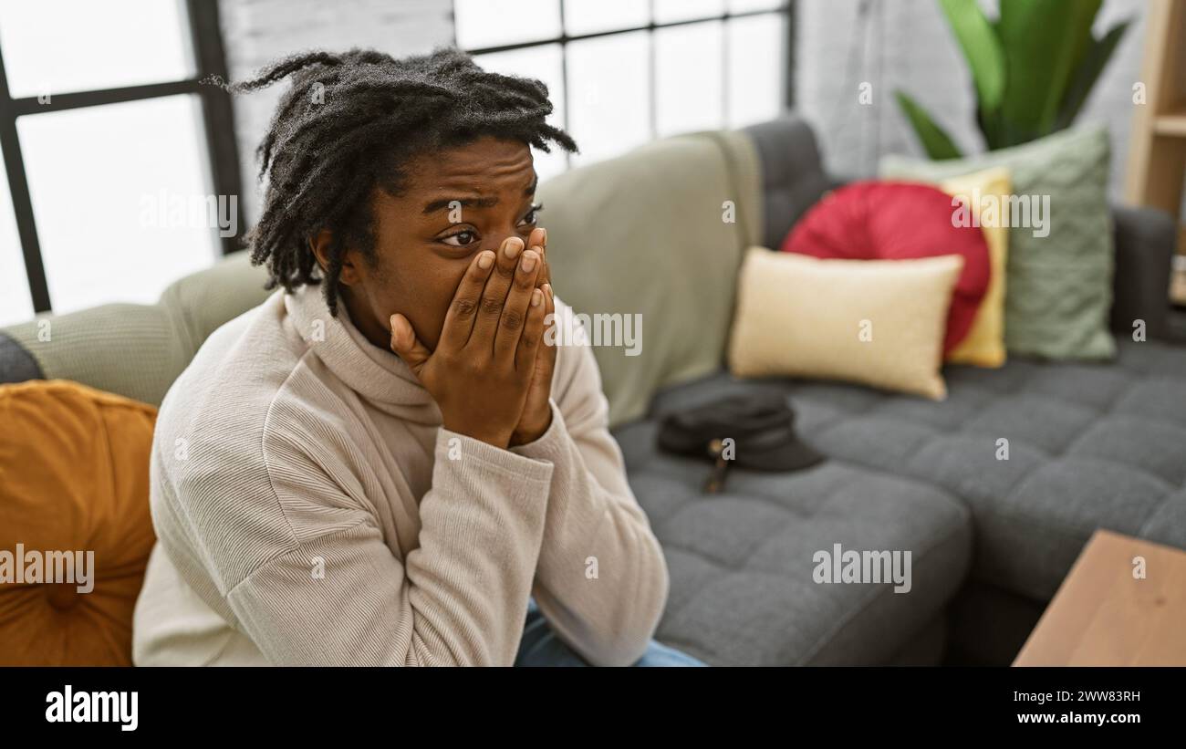 Surprised young black woman with dreadlocks sitting on a sofa in a cozy ...