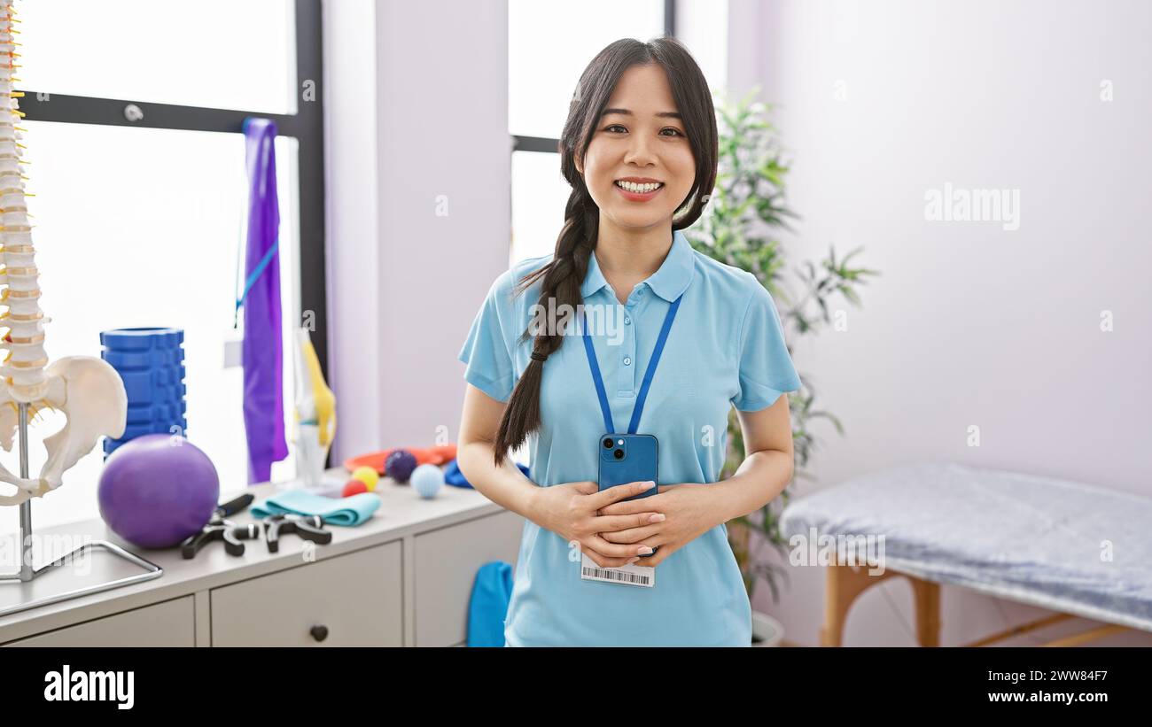 Asian woman therapist with phone smiling in a physiotherapy clinic ...