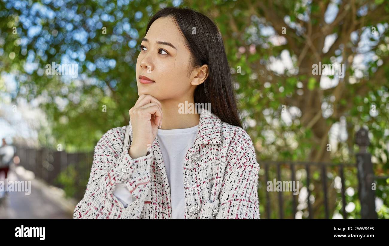 Thoughtful asian woman in casual attire posing outdoors in a lush city ...