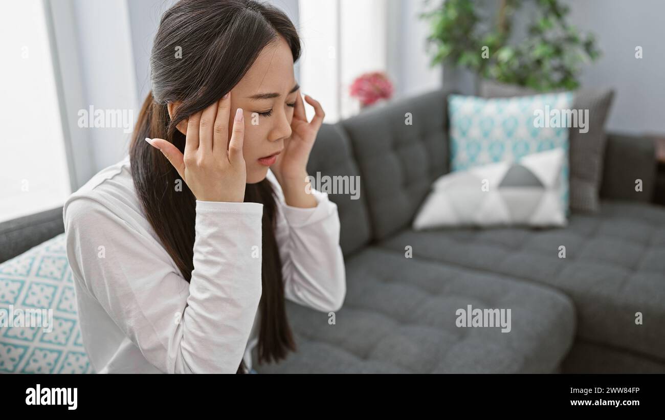 A stressed chinese woman with a headache at home, sitting on a couch in ...