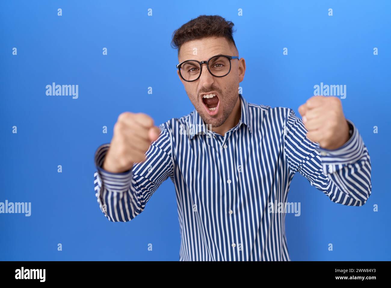 Hispanic man with beard wearing glasses angry and mad raising fists ...
