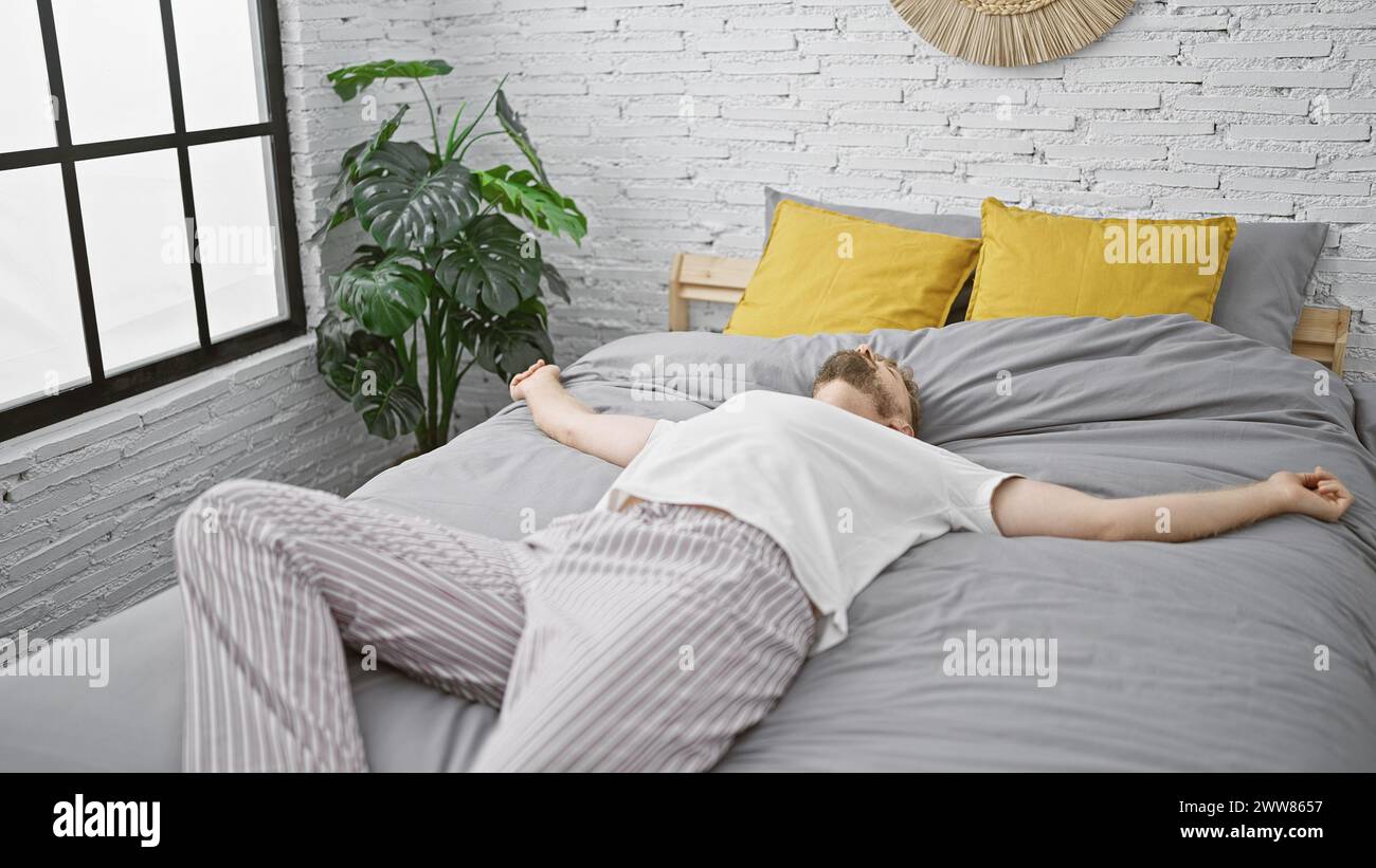 A young caucasian man with a beard is sprawled out on a bed in a modern bedroom, expressing ...