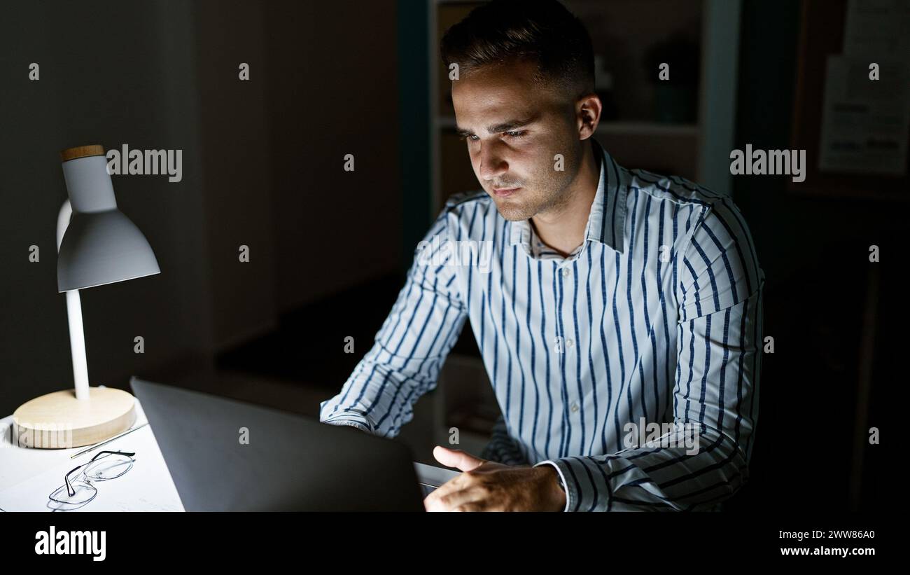 Young man working late in the office, illuminated by a desk lamp, using ...
