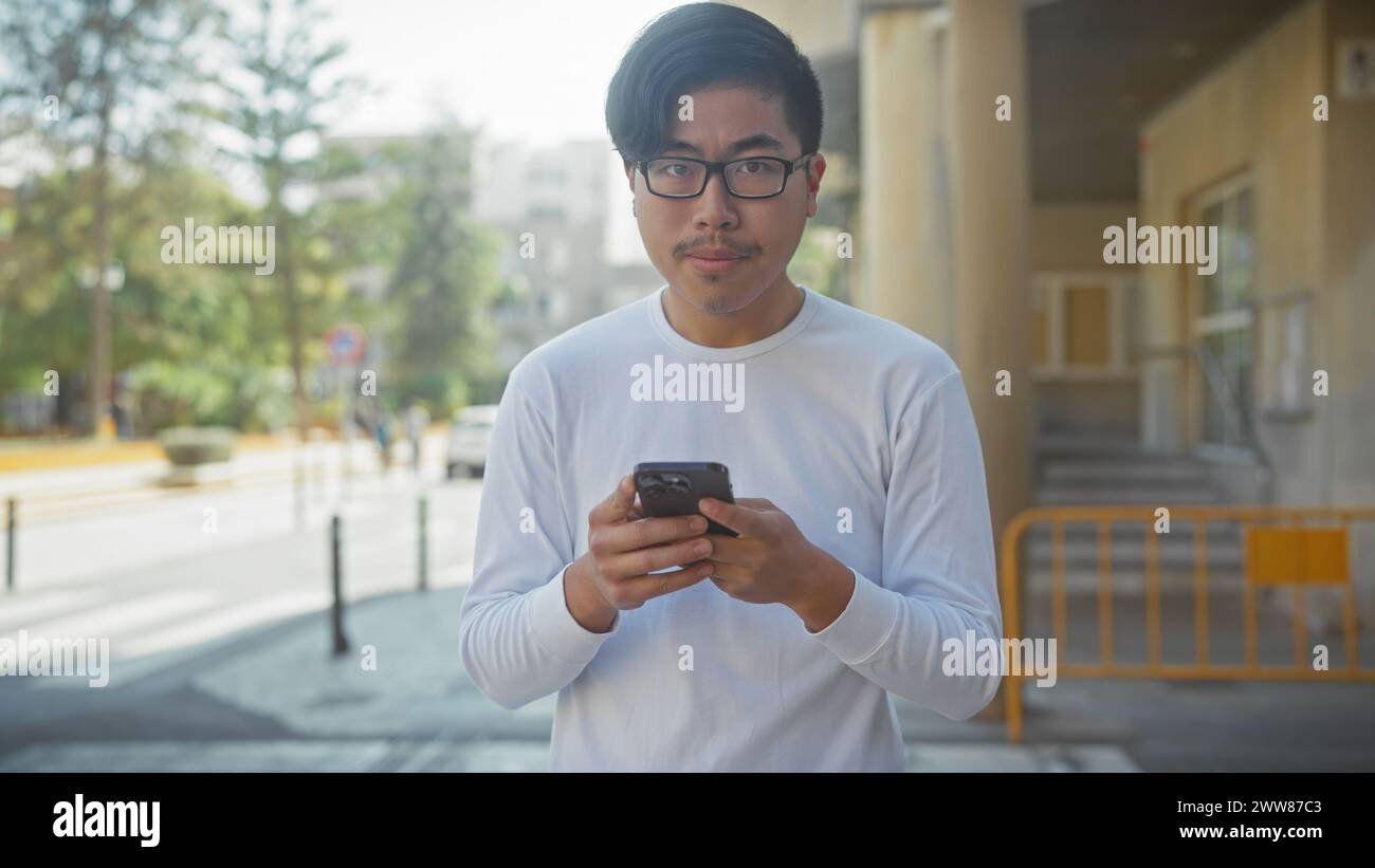 A young asian man uses a smartphone while standing on a city street ...