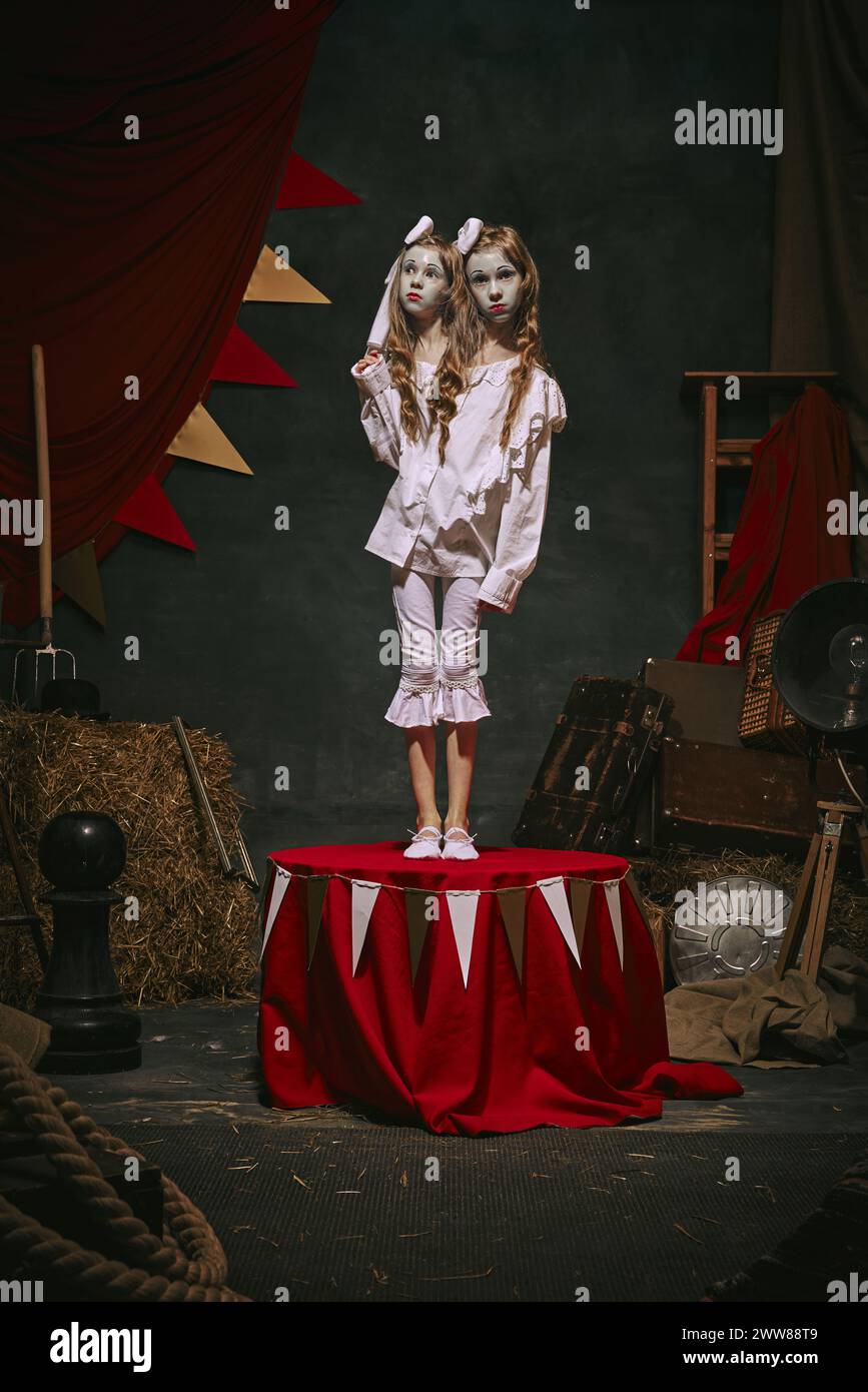 Siamese twin girls in white vintage costumes with makeup stand on stage over dark retro circus ...