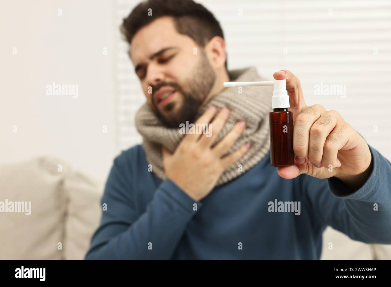 Young man with scarf holding throat spray indoors, selective focus ...