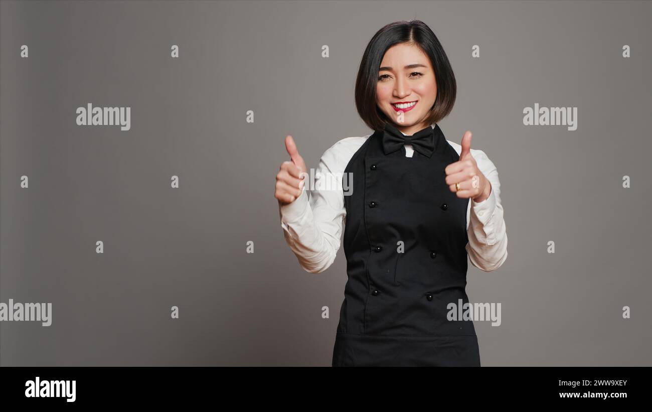 Smiling waitress giving thumbs up sign in front of camera, working in a ...