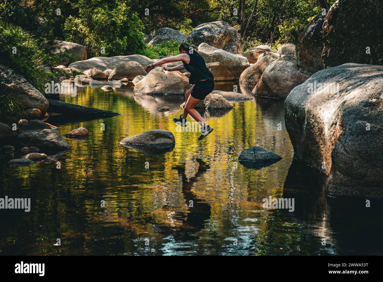 Adventurous hiker making a bold jump over a calm river with boulders ...