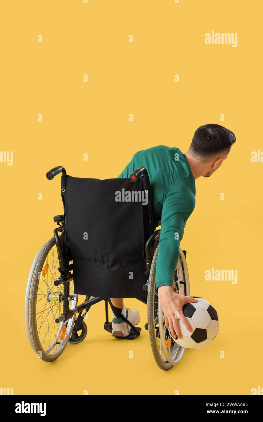 Male footballer in wheelchair with ball on yellow background, back view ...