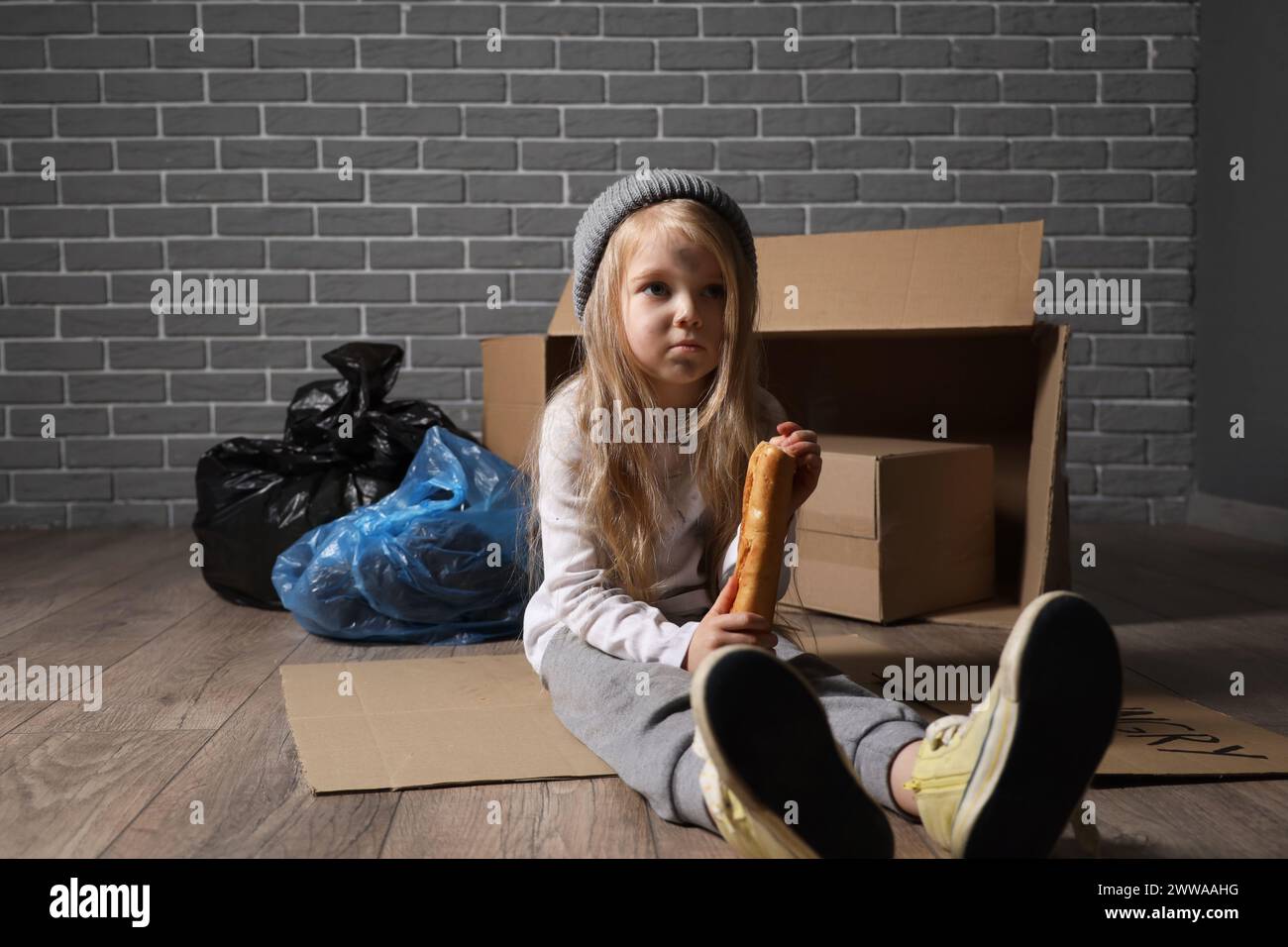 Homeless little girl eating bread near trash against grey brick wall ...