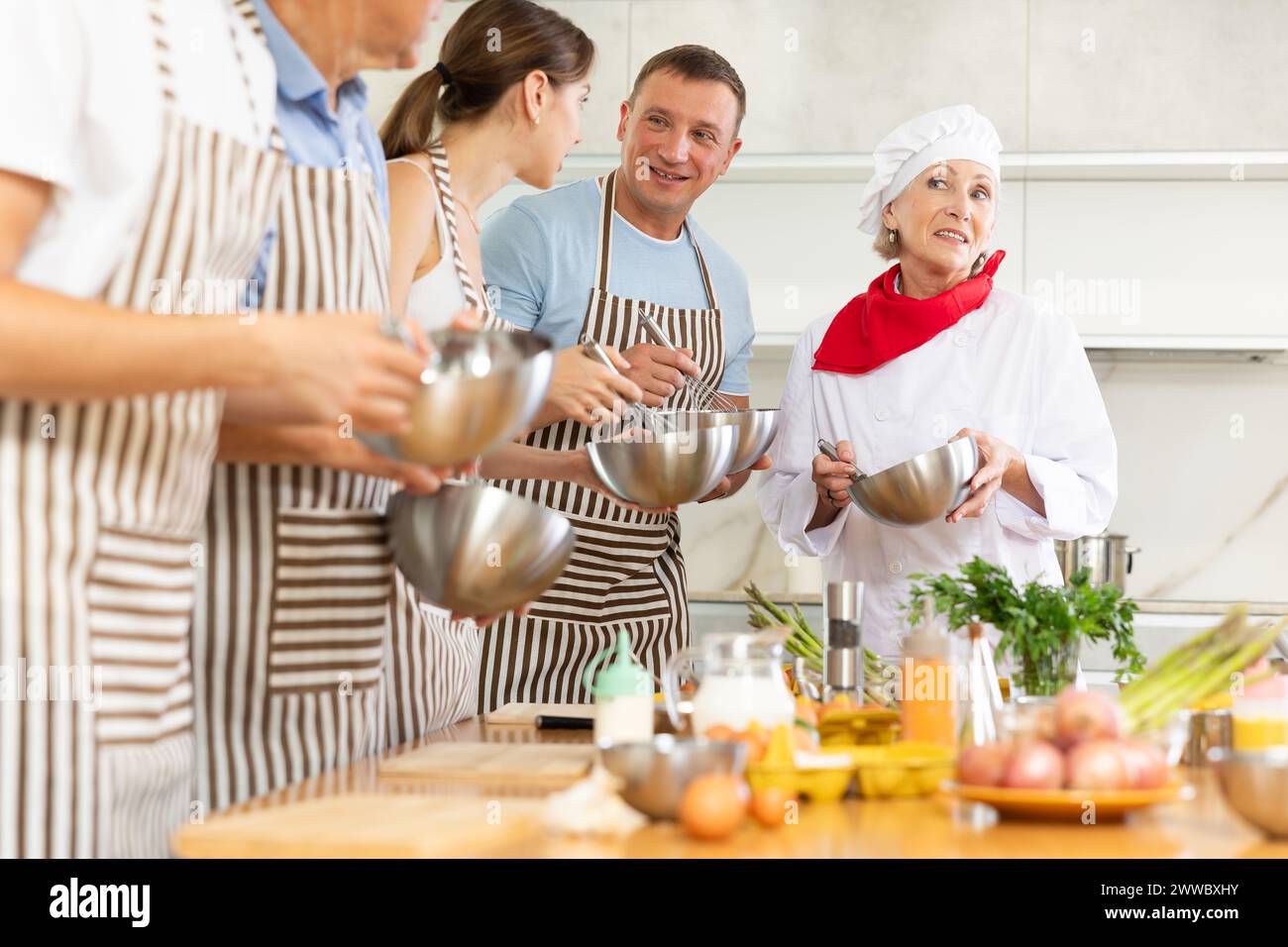 Smiling old woman chef of cooking course teaching attendees how to mix ...