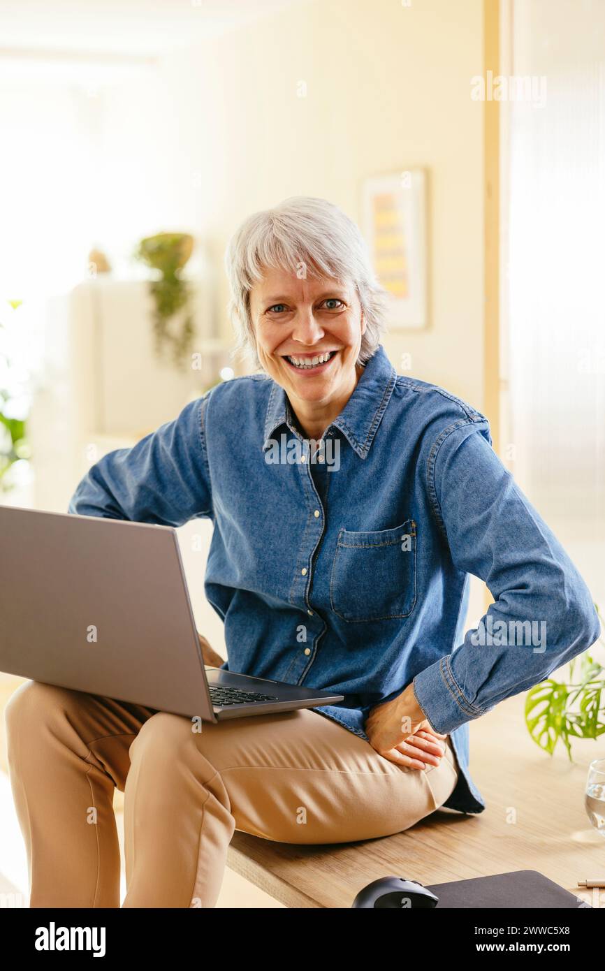 Happy freelancer sitting on table with arms akimbo at home office Stock ...