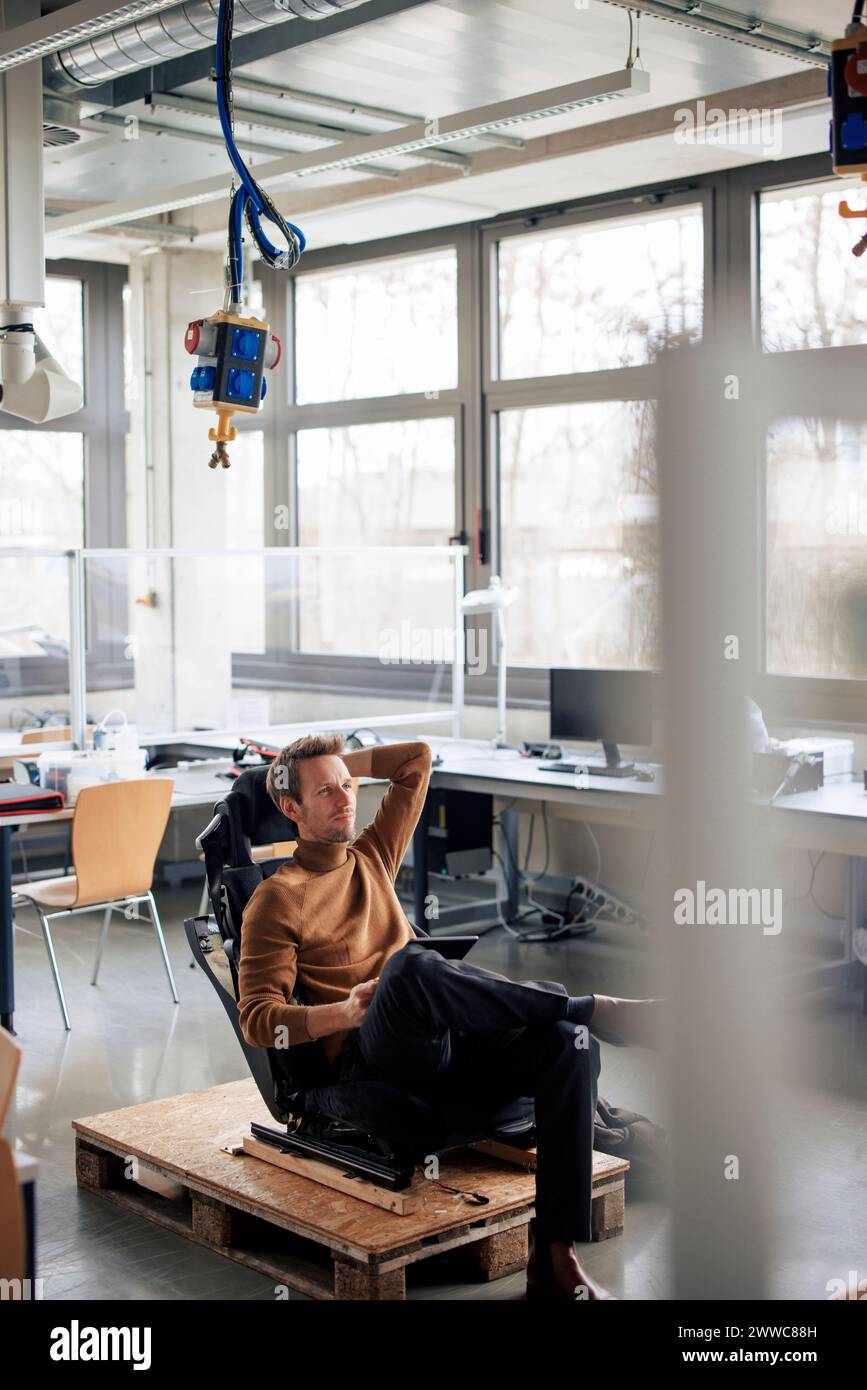 Relaxed engineer sitting with hand behind head on chair at workshop ...