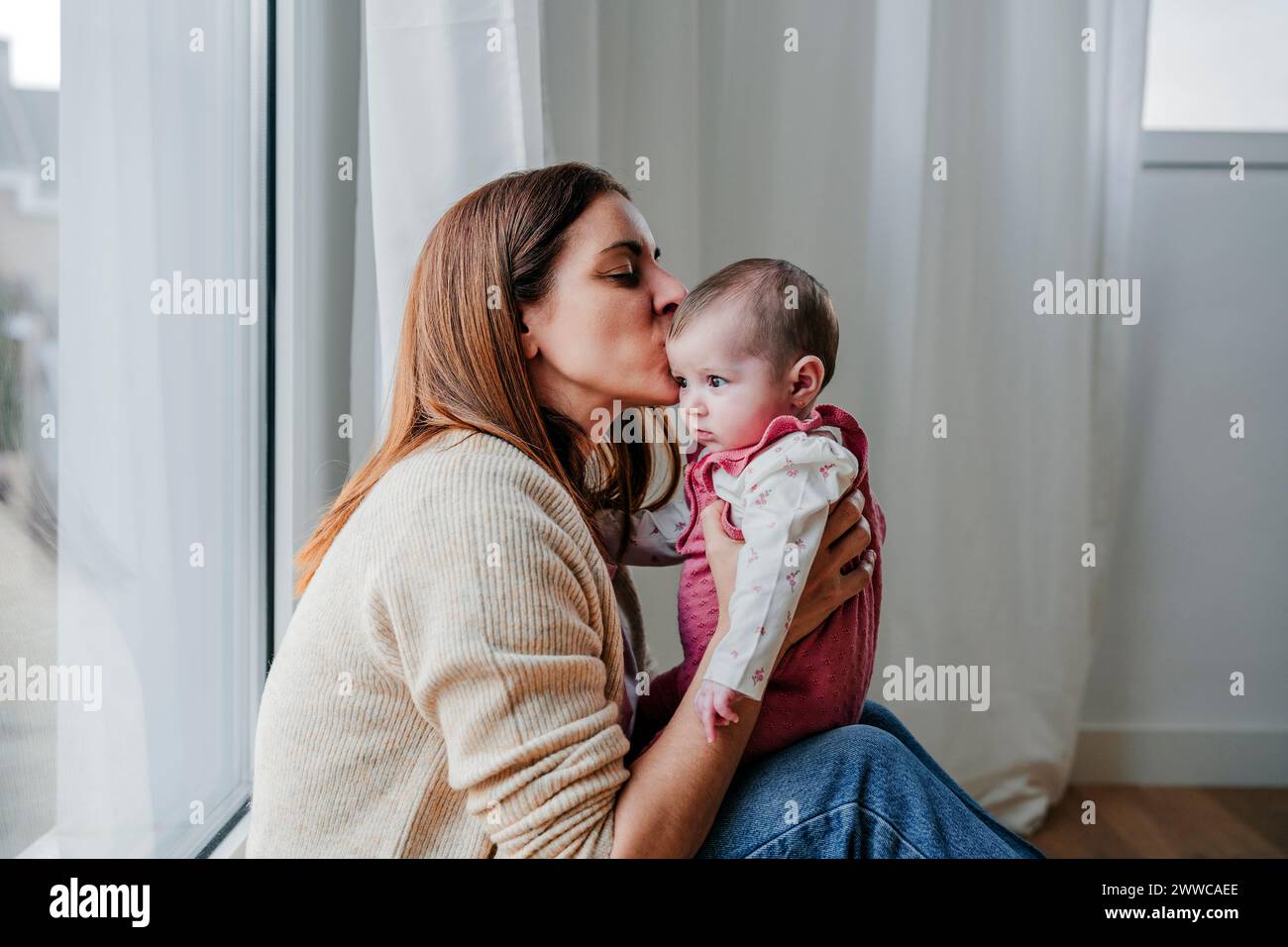 Mother kissing baby daughter sitting in front of window Stock Photo - Alamy