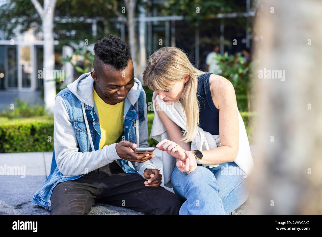 Curious multiracial friends sharing smart phone sitting on bench Stock ...