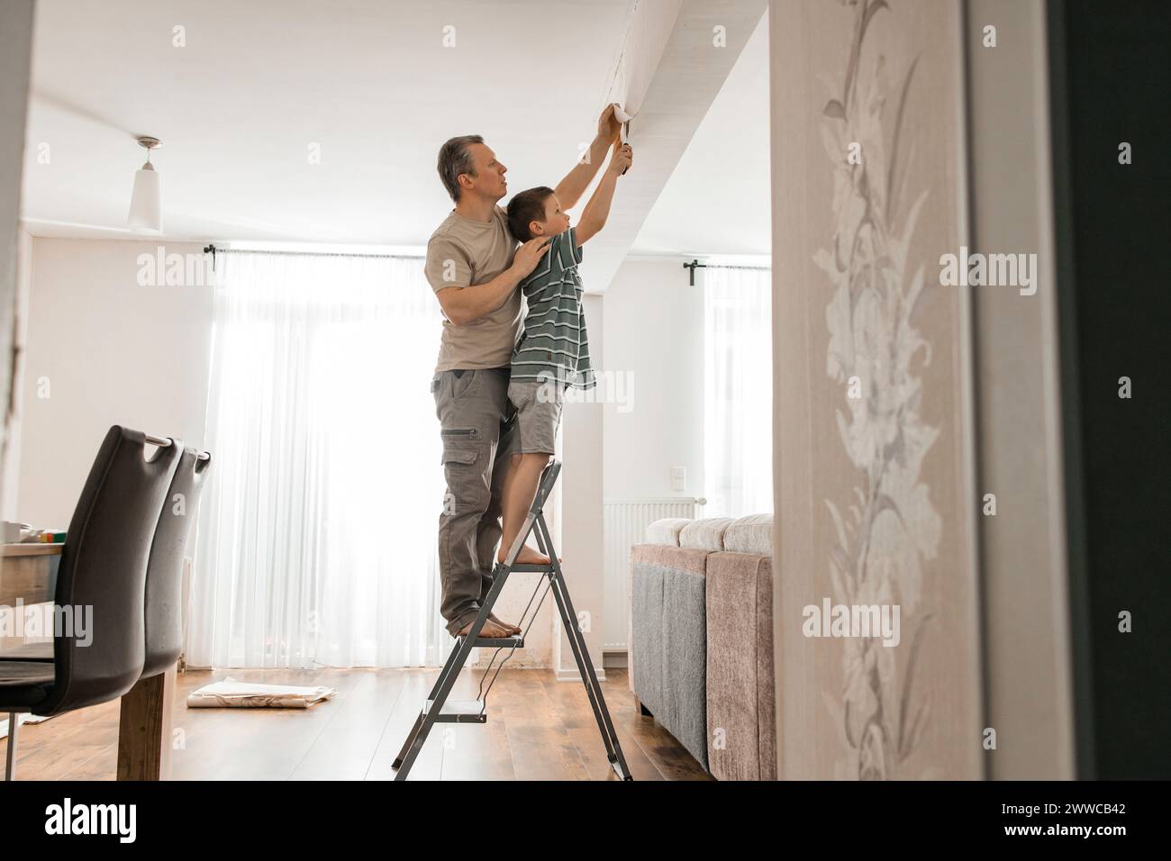 Son and father peeling old wallpaper standing on step ladder at home ...