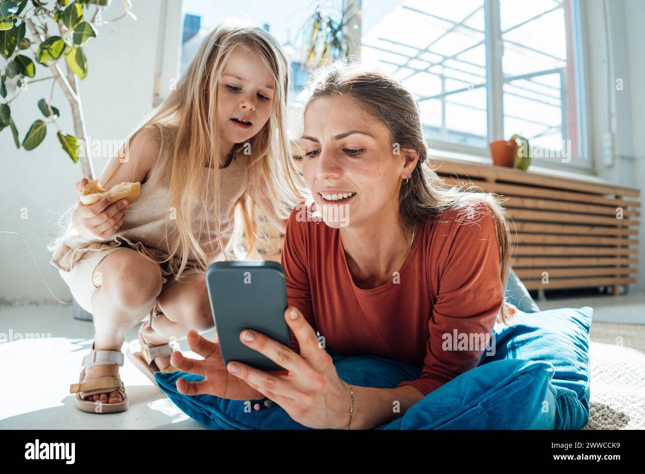 Smiling woman using smart phone with daughter holding snack at home ...