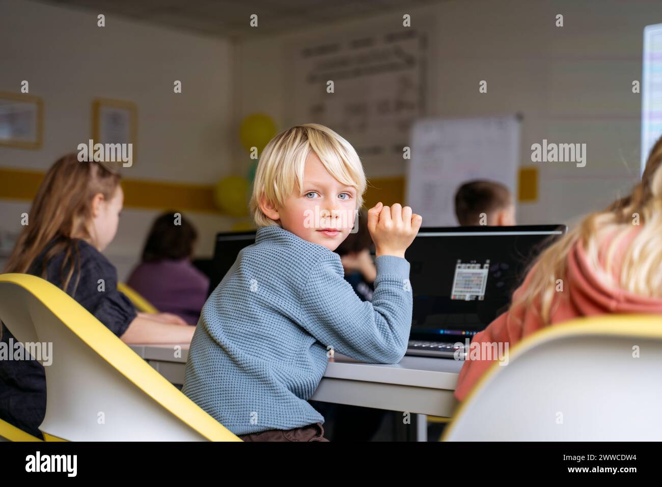 Blond boy looking back over shoulder sitting with laptop at desk in ...