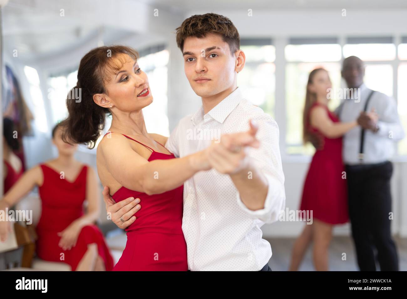 Pretty young woman dancing waltz with partner at group of multinational people in ballroom ...