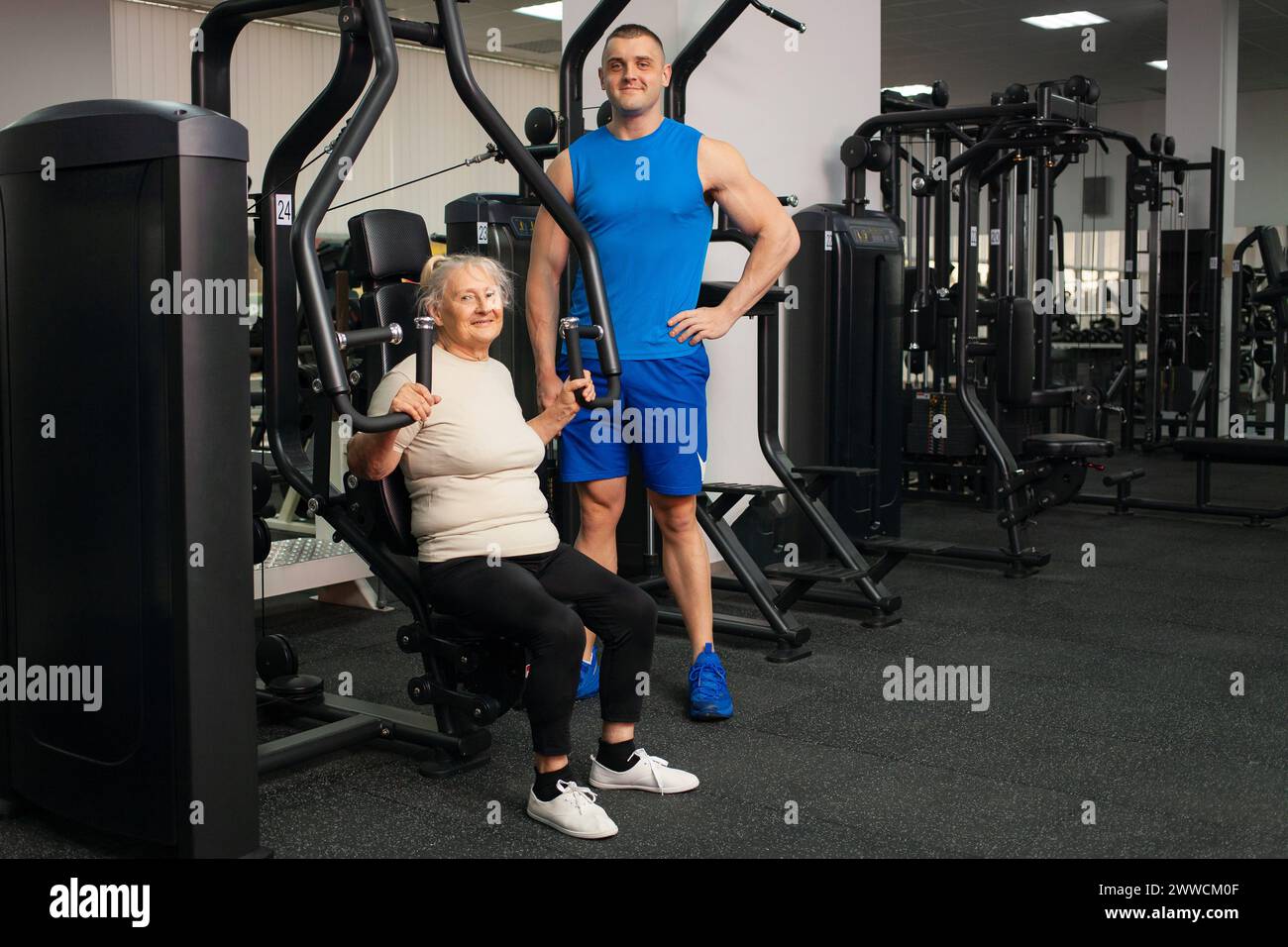 A handsome young coach man helps an old woman train sports in the gym ...