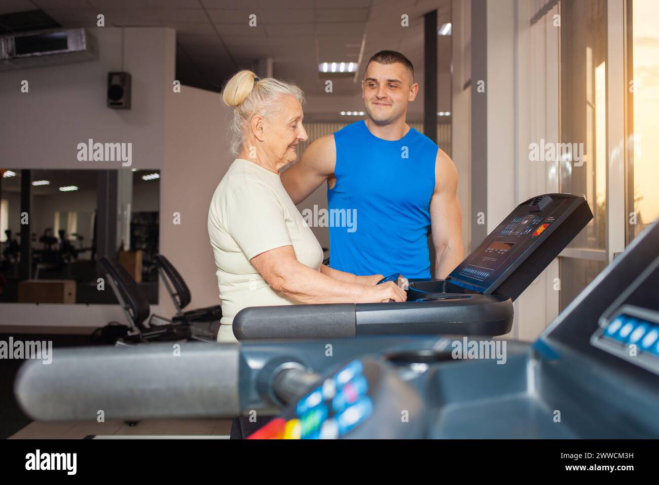 A young handsome coach shows an old woman how to manage a treadmill ...