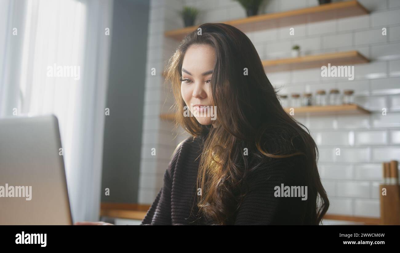 Asian woman sitting in the kitchen typing on laptop chatting with ...