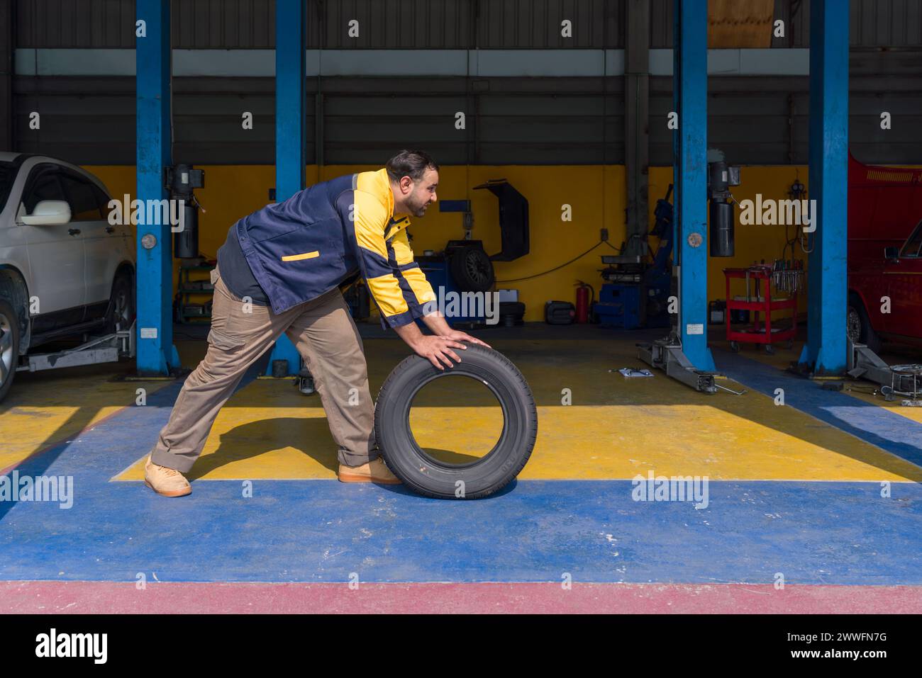 A mechanic in uniform rolling a tire across workshop or garage floor ...