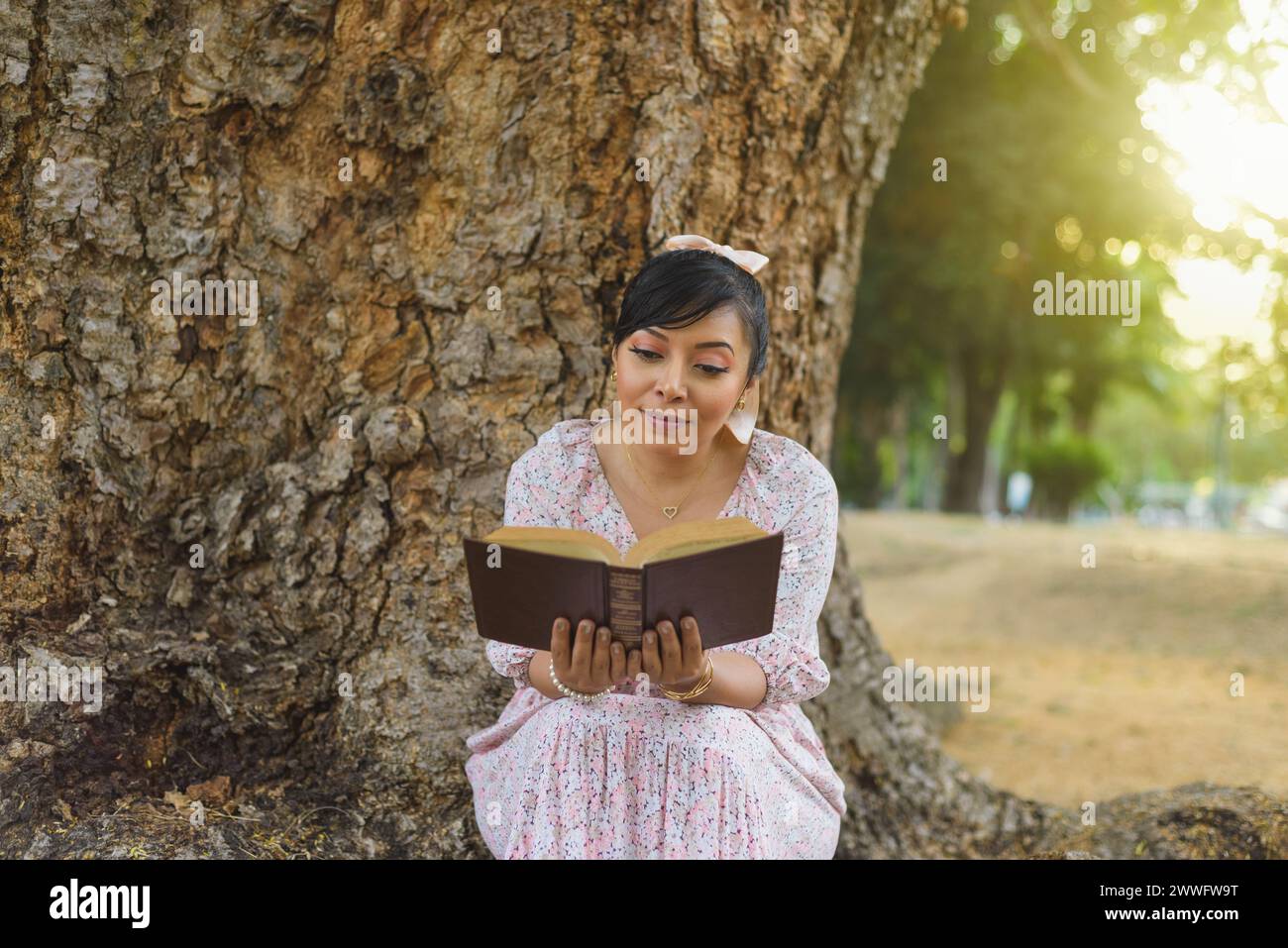 Woman seated next to a tree trunk in a park reading a book. World book ...