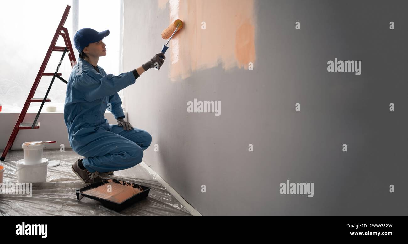 female painter in overalls with cap and gloves painting a wall with ...