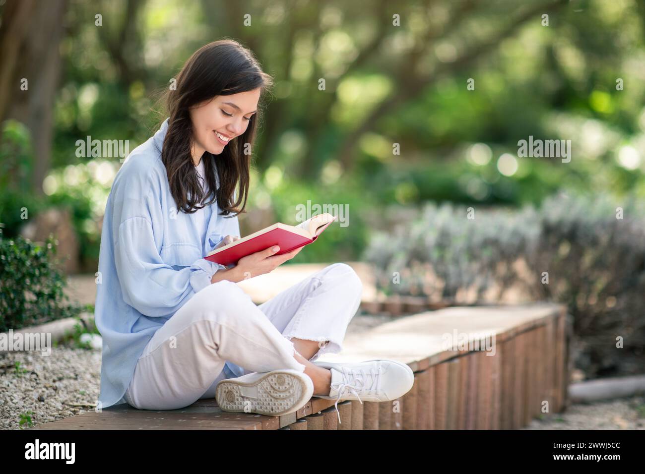 Serene young woman enjoying reading a red book, sitting cross-legged on ...