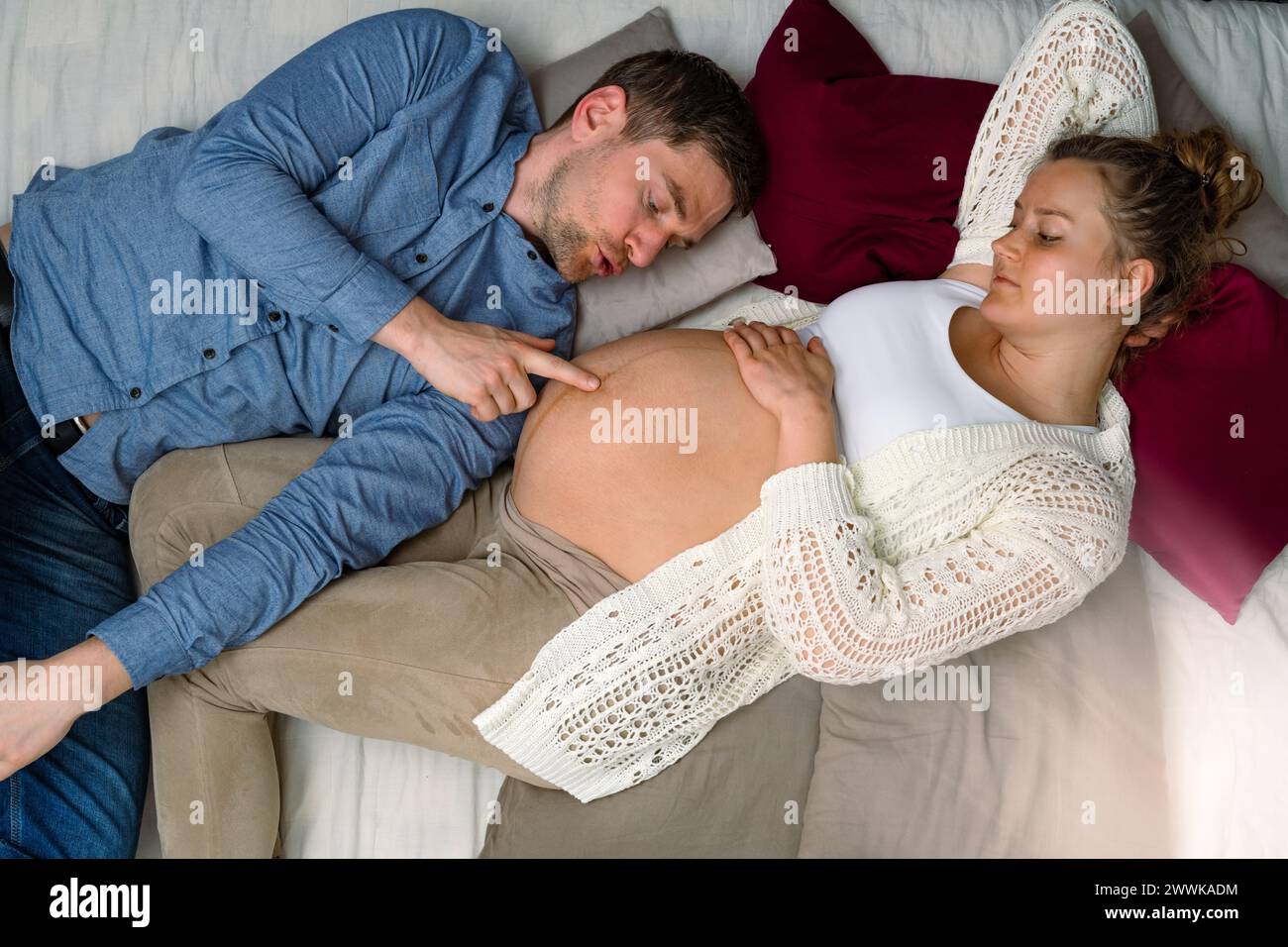 Description: Expectant parents lie in bed and the father gently pokes ...