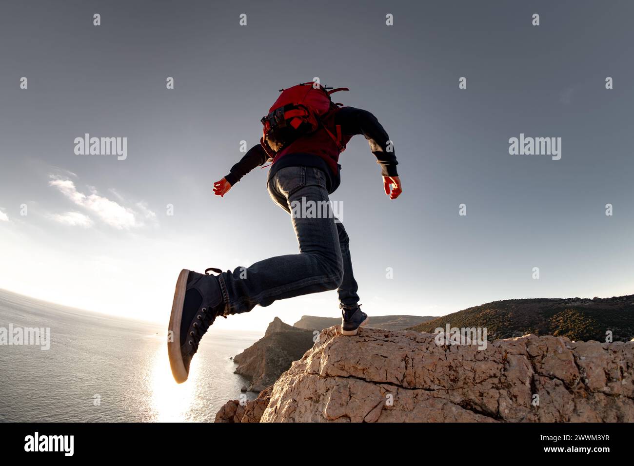 Active hiker with backpack jumps to big rock against sunset sky and sea ...