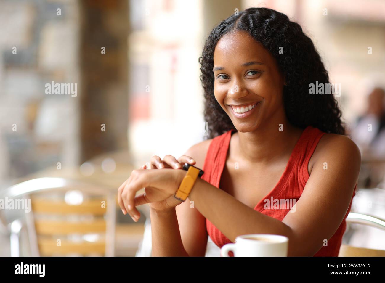 Happy black woman with smartwatch looks at you in a restaurant terrace ...