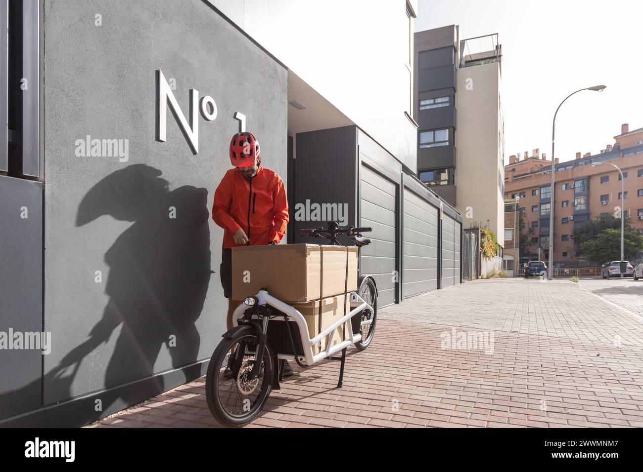 young courier with red clothing and helmet riding cargo bike arriving at the shipping ...