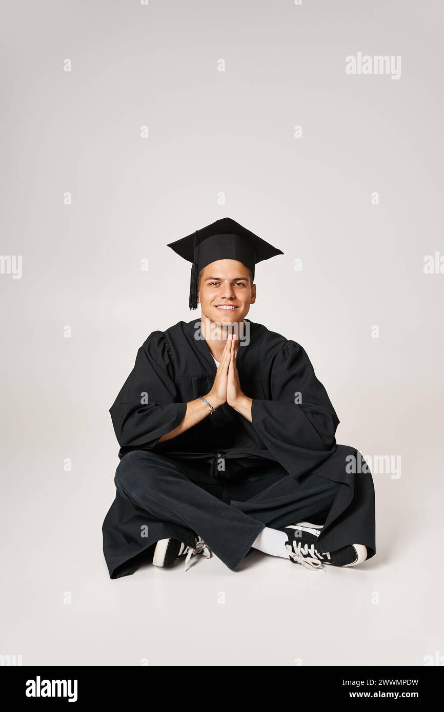 attractive young guy in graduate gown and cap sitting and folding hands ...