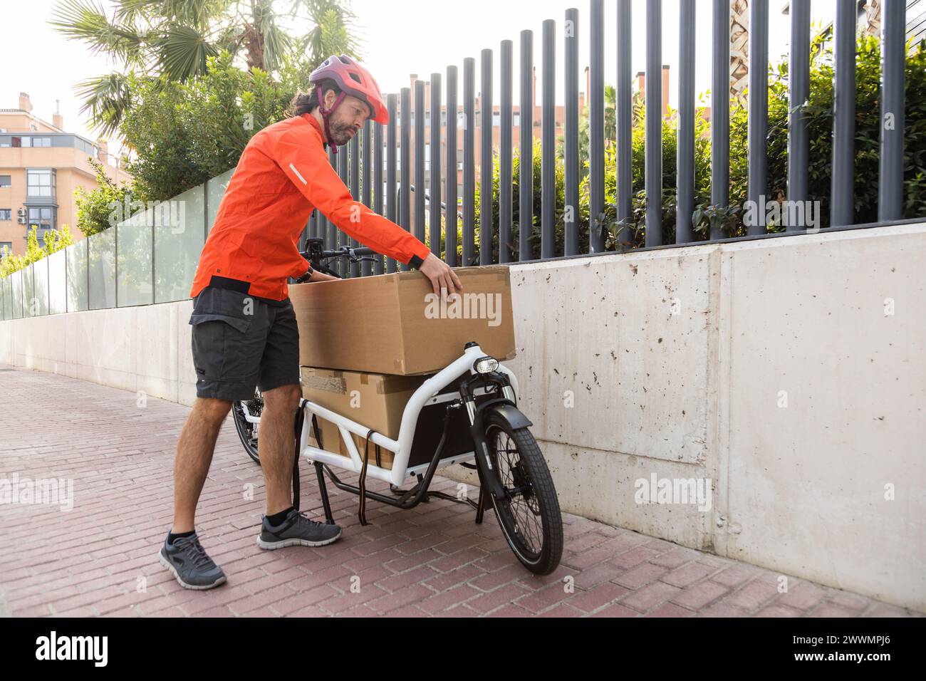 young courier with red clothing and helmet riding cargo bike arriving at the shipping ...