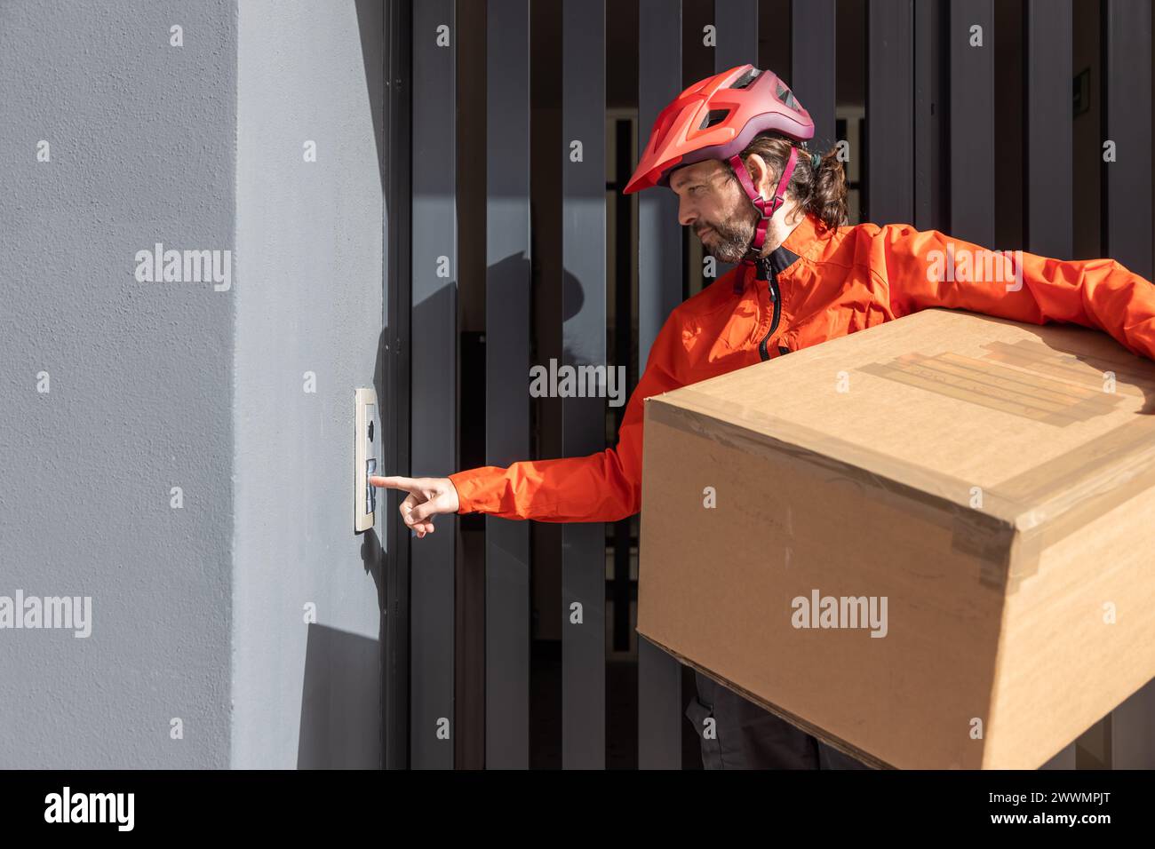 young courier with red clothing and helmet riding cargo bike arriving at the shipping ...