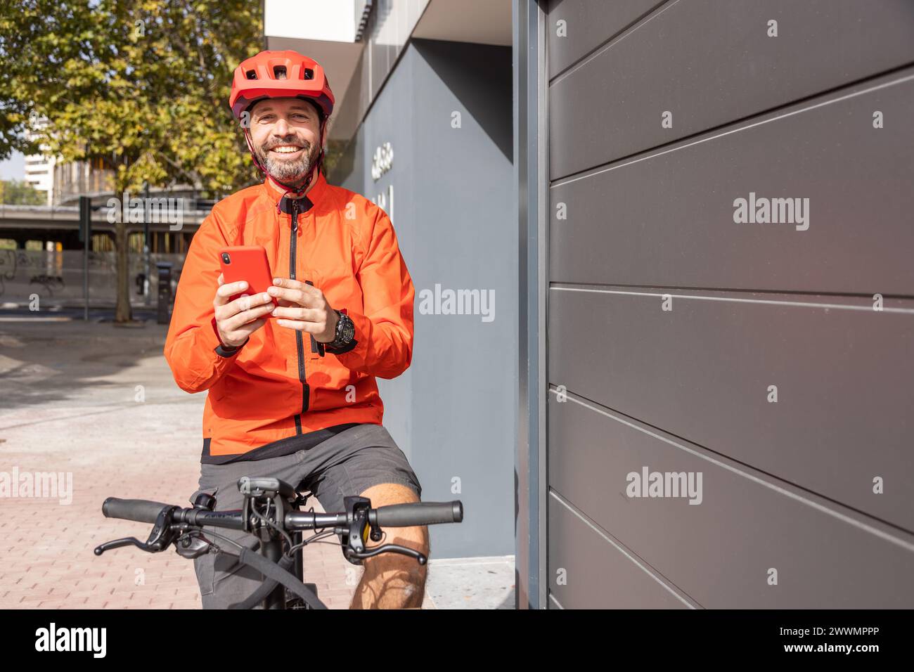 young courier with red clothing and helmet riding cargo bike arriving at the shipping ...