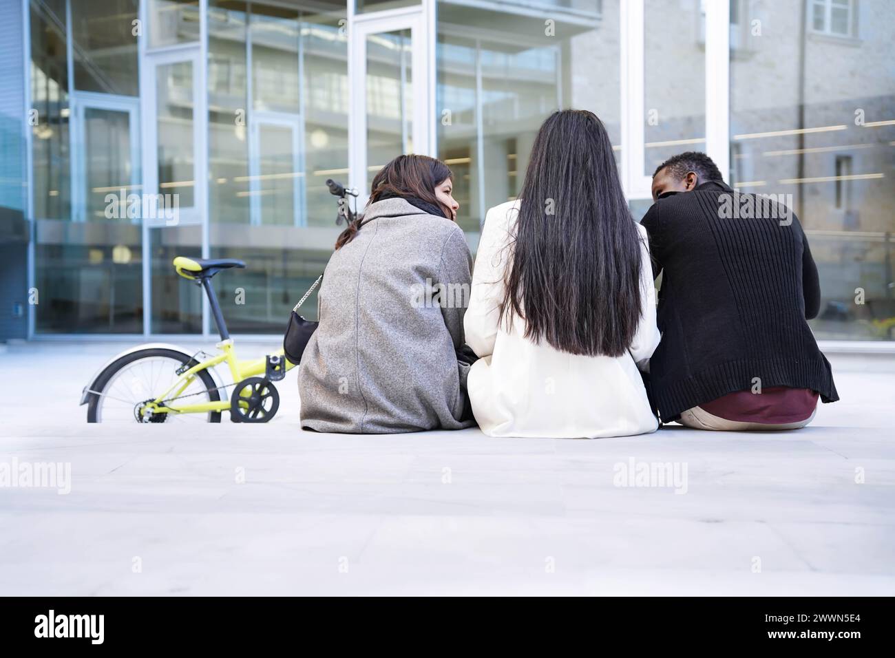 Three multiracial people sitting on the ground in front of a office ...