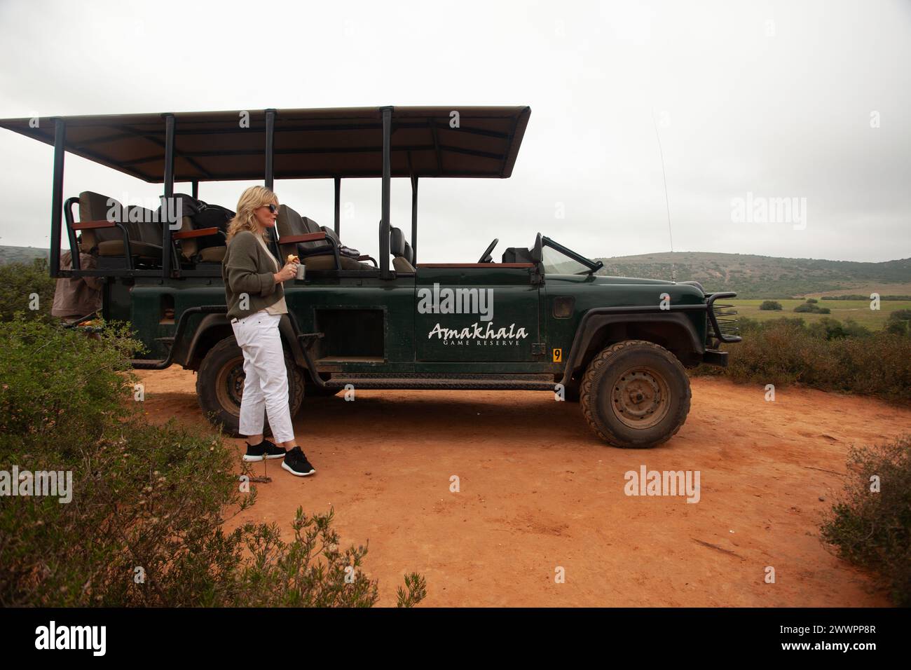 Blonde Woman In White Trousers Drinks A Beverage Standing Beside A Safari Jeep In The Amakhala
