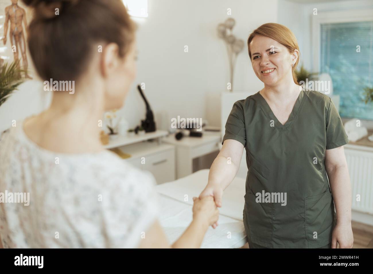 Healthcare time. smiling female medical massage therapist in massage cabinet with client shaking ...