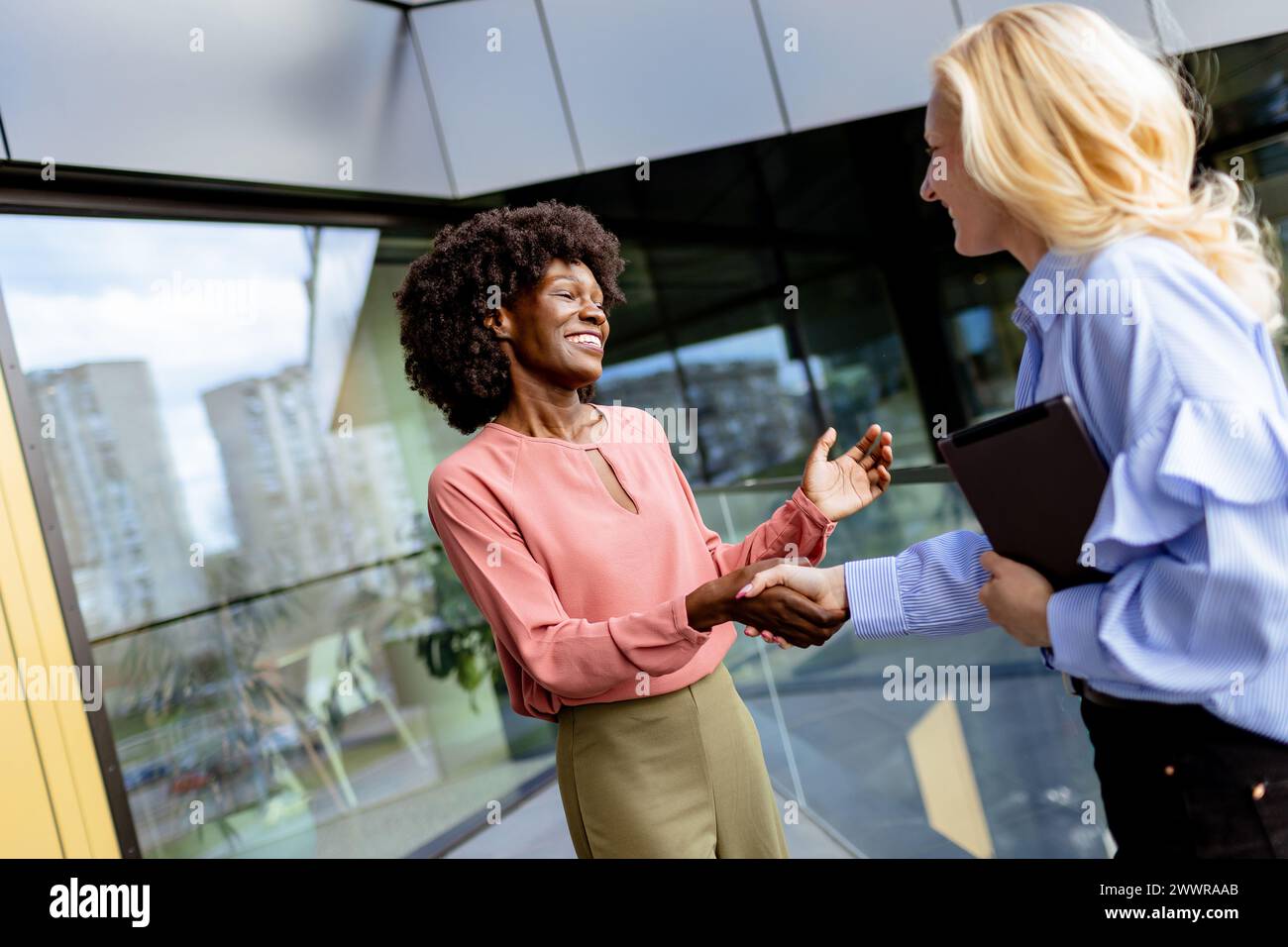 Two colleagues laugh together, one holding a tablet in a sunny, glass ...